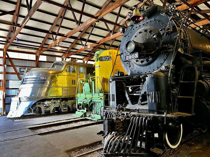 Three legends under one roof: a gleaming silver streamlined engine, a vibrant yellow locomotive, and a powerful black steam engine stand ready, like magnificent time machines waiting to tell tales of railroading's golden eras.