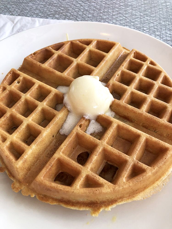 Golden waffle perfection with a melting pat of butter—architectural breakfast brilliance with those perfect squares collecting just the right amount of syrup.