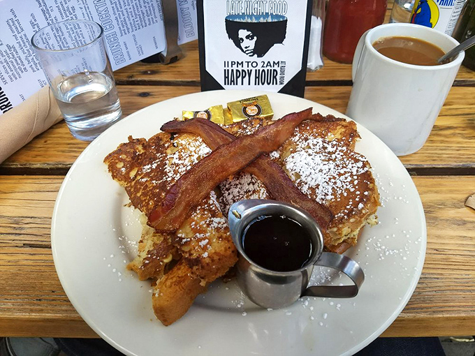 Breakfast alchemy: French toast crowned with powdered sugar and bacon crosses that transforms ordinary mornings into celebrations. The tiny pitcher of maple syrup is basically liquid gold.