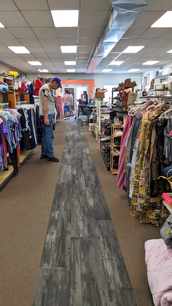 A narrow grey-brick walkway leads through the center of a bustling thrift store, guiding shoppers past racks of clothing and vintage finds.