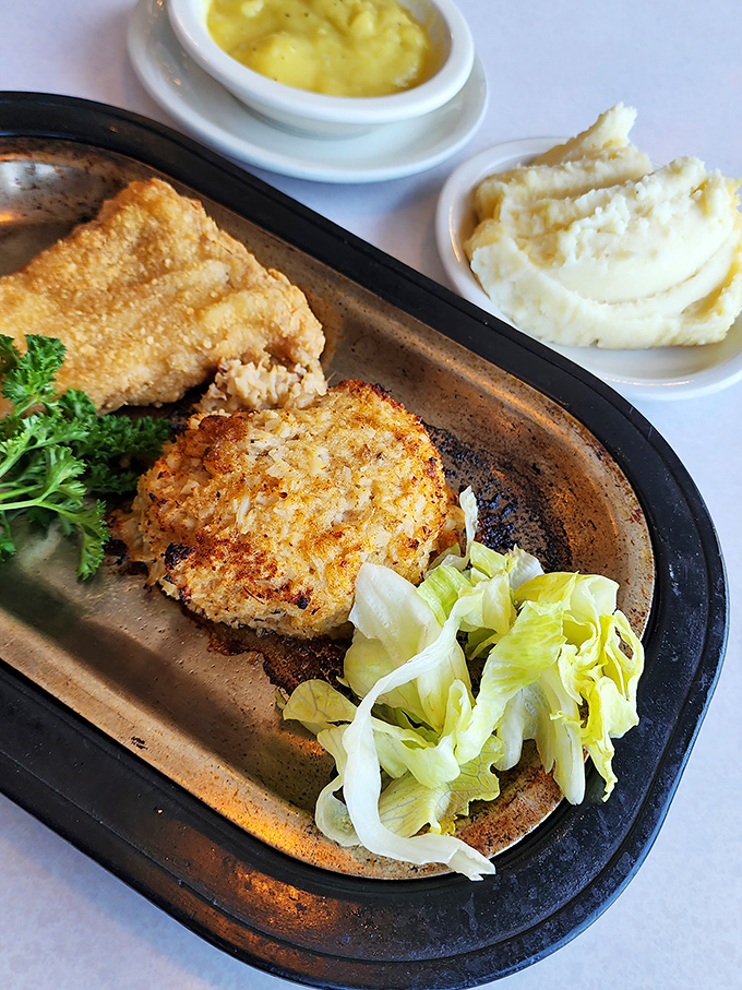 The crab cake sits proudly on its sizzling throne, wearing a golden crust like armor protecting treasures within.