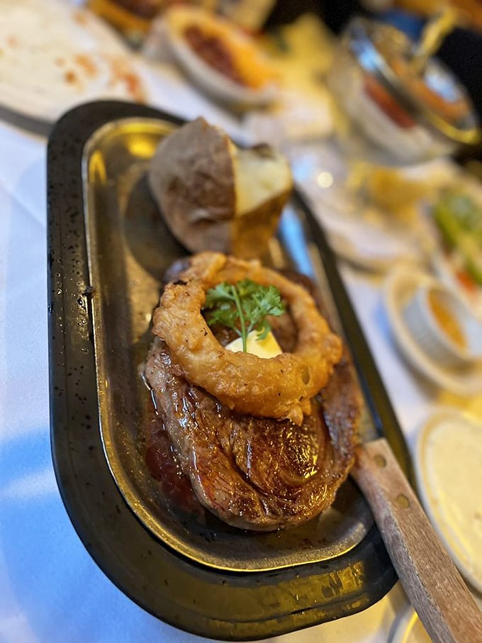 The ribeye arrives like a meaty masterpiece on its metal throne, topped with an onion ring portal to flavor heaven that would make carnivores weep with joy.