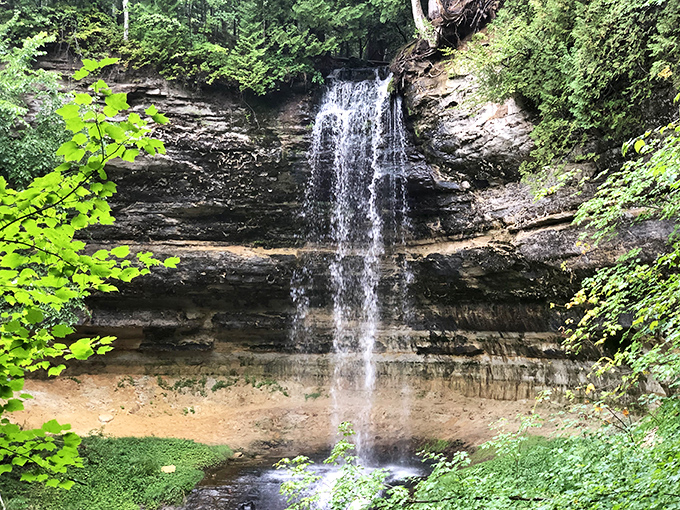 Water cascades in a delicate ribbon, creating its own soundtrack as it tumbles down layered rock faces into a serene pool below.