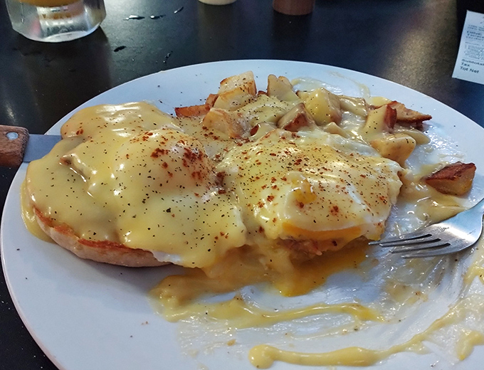 Eggs Benedict done diner-style: perfectly poached eggs lounging on English muffins while hollandaise sauce cascades down like a yellow waterfall of happiness.