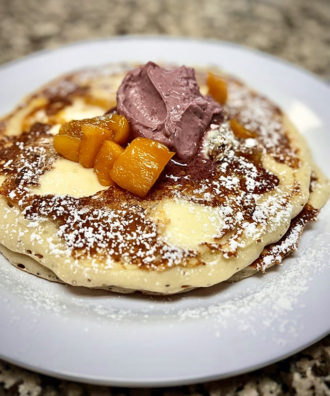 These pancakes don't need Instagram filters. Golden discs topped with fruit, powdered sugar, and what appears to be chocolate butter&mdash;breakfast that doubles as emotional support.
