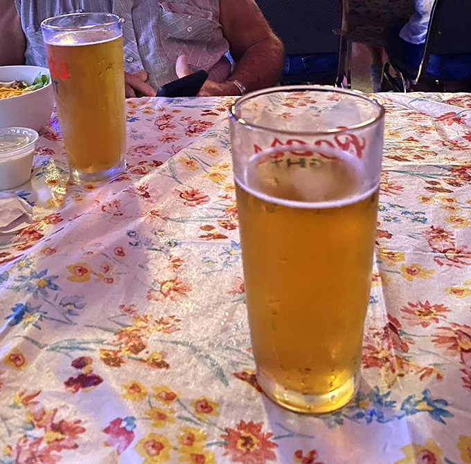 Cold beer in tall glasses—nature's perfect companion to smoked meats. The floral tablecloth adds a touch of grandma's kitchen to your BBQ adventure.
