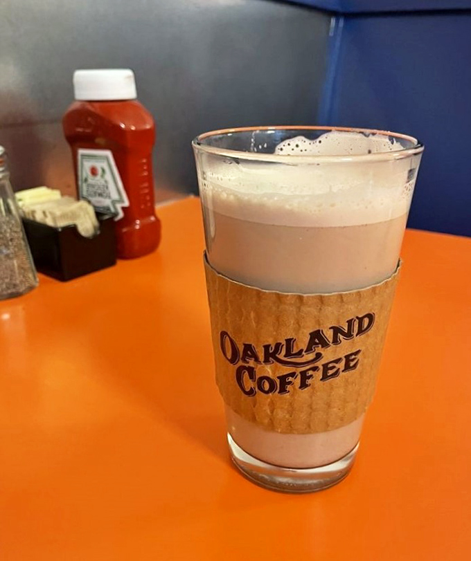 Oakland Coffee in a glass that says "I'm not just caffeine, I'm an experience." The cardboard sleeve isn't just practical&mdash;it's a warm handshake.