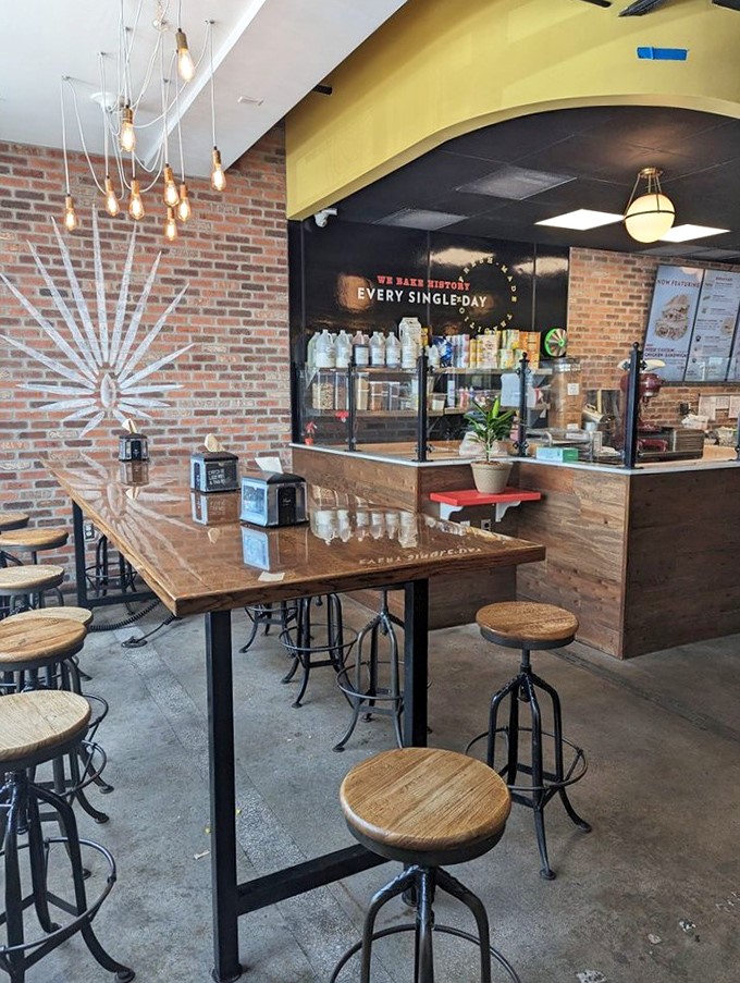 The community table where strangers become friends over shared bread. Edison would approve of those hanging bulbs illuminating culinary conversations.