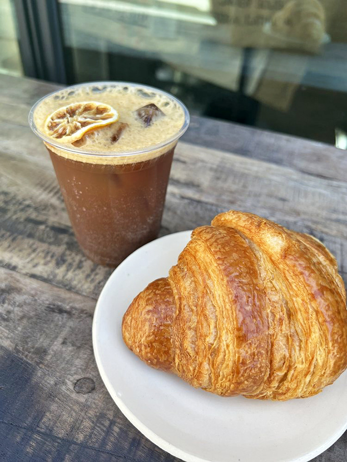 The holy breakfast trinity: golden croissant, artfully crafted cold brew, and weathered wood table&mdash;simple pleasures that somehow feel revolutionary in their perfection.