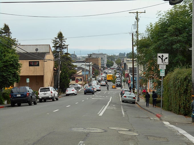This photo captures a lively street scene with a mix of cars, cyclists, and pedestrians set against a backdrop of small businesses and distant coastal views.