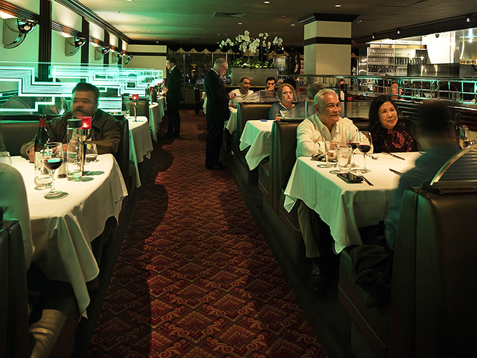 The restaurant hums with the universal language of good food and conversation, as diners dive into their steakhouse experience under atmospheric green lighting.