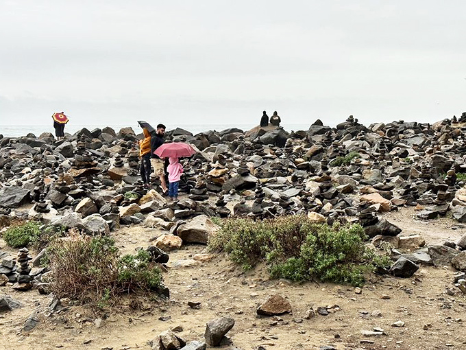 Explorers navigate a rocky shoreline under moody skies. The pink umbrella adds a touch of whimsy to this geological playground where everyone becomes a rock stacker.