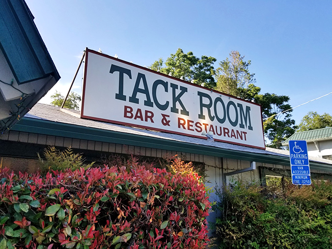 The unassuming exterior of Tack Room Bar & Restaurant, where vibrant foliage frames a sign that promises more than it shows&mdash;culinary magic awaits inside.