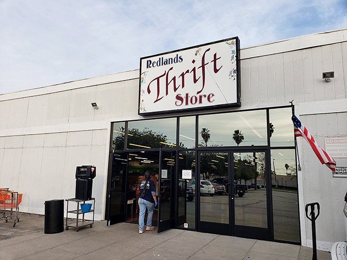 The unassuming exterior of Redlands Thrift Store, where treasures hide behind a simple sign and an American flag waves a welcome to bargain hunters.