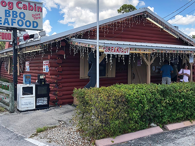 The rustic red exterior of Log Cabin BBQ stands as a beacon of comfort food in LaBelle, where Christmas lights twinkle year-round and promises of BBQ and seafood beckon hungry travelers.