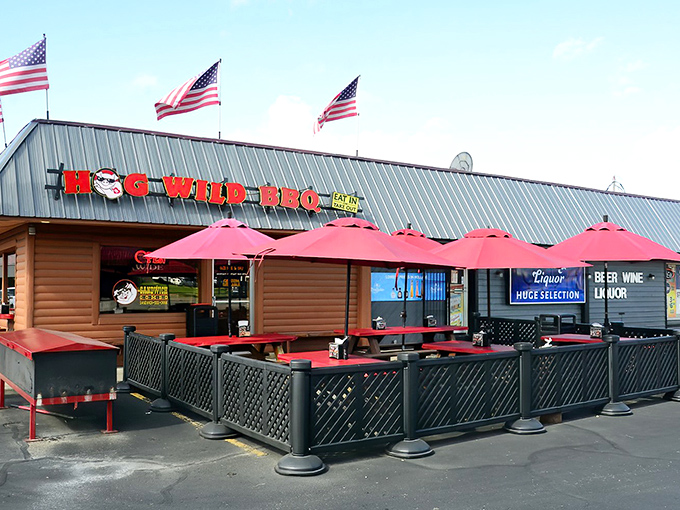 American flags flutter above Hog Wild's rustic exterior like patriotic smoke signals, beckoning hungry travelers with the promise of barbecue bliss beneath those cheerful red umbrellas.