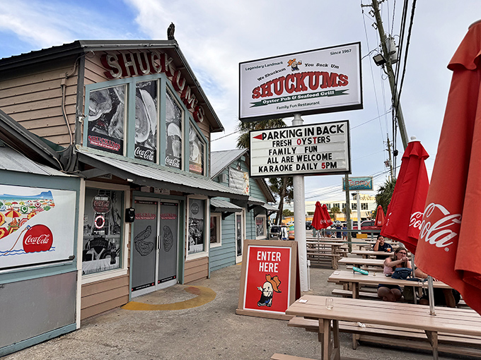 The weathered blue exterior of Shuckums stands like a salty sea captain, promising treasures of the deep within. Those red umbrellas beckon like maritime flags signaling "good eats ahead."