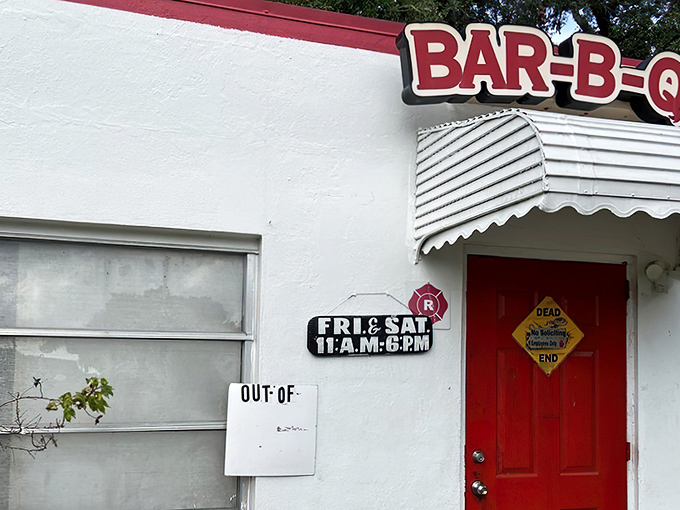 Simplicity at its finest: a modest white building with red trim, shaded by Florida oaks, promising smoky delights that need no fancy facade.