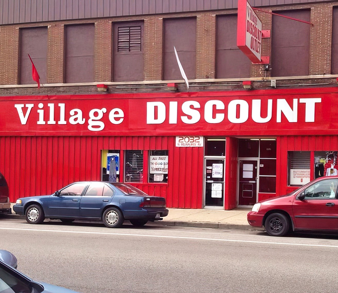 The crimson facade of Village Discount stands like a beacon for bargain hunters, its bold white lettering promising treasures within those no-frills walls.