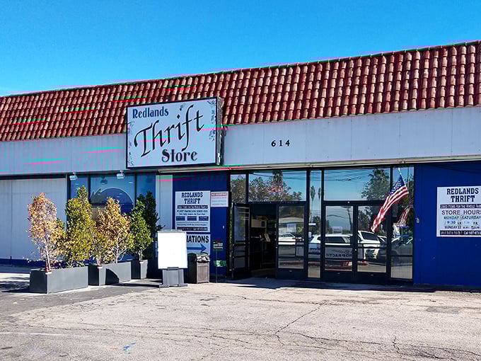 The unassuming exterior of Redlands Thrift Store, where treasures hide behind a simple sign and an American flag waves a welcome to bargain hunters.