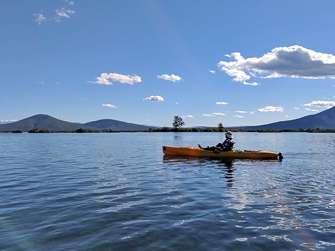 Solitude perfected! A lone kayaker glides across mirror-like waters, with mountains creating nature's version of surround sound scenery.