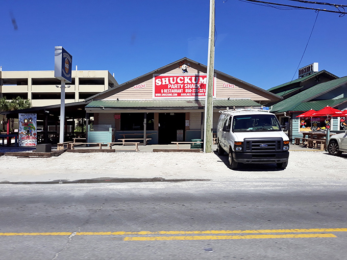 The weathered blue exterior of Shuckums stands like a salty sea captain, promising treasures of the deep within. Those red umbrellas beckon like maritime flags signaling "good eats ahead."