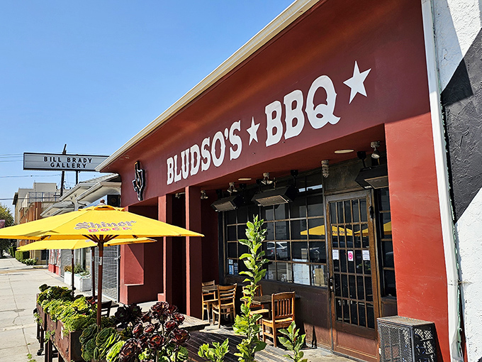 The red storefront of Bludso's BBQ stands like a beacon of hope for hungry travelers, promising smoky salvation under those bright yellow umbrellas.