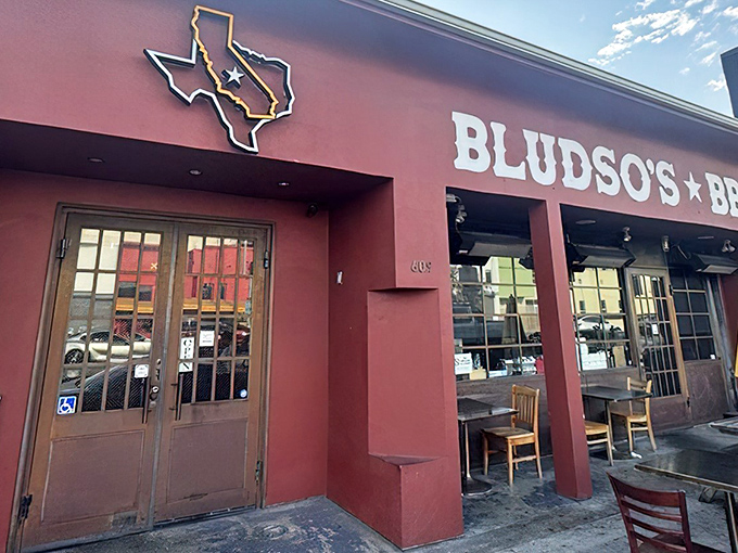 The red storefront of Bludso's BBQ stands like a beacon of hope for hungry travelers, promising smoky salvation under those bright yellow umbrellas.
