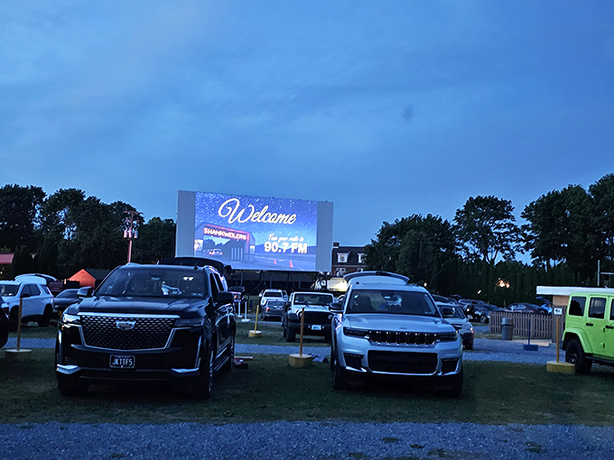 Modern cars line up facing the illuminated screen at dusk, where technology meets nostalgia under Pennsylvania's darkening sky.
