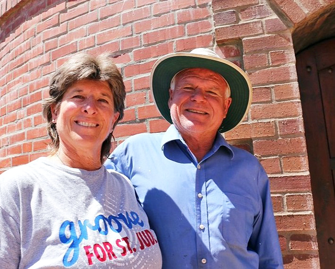 The image shows two people standing by a brick wall, smiling in the sunshine, embodying the warm welcome visitors receive at this unique California attraction.
