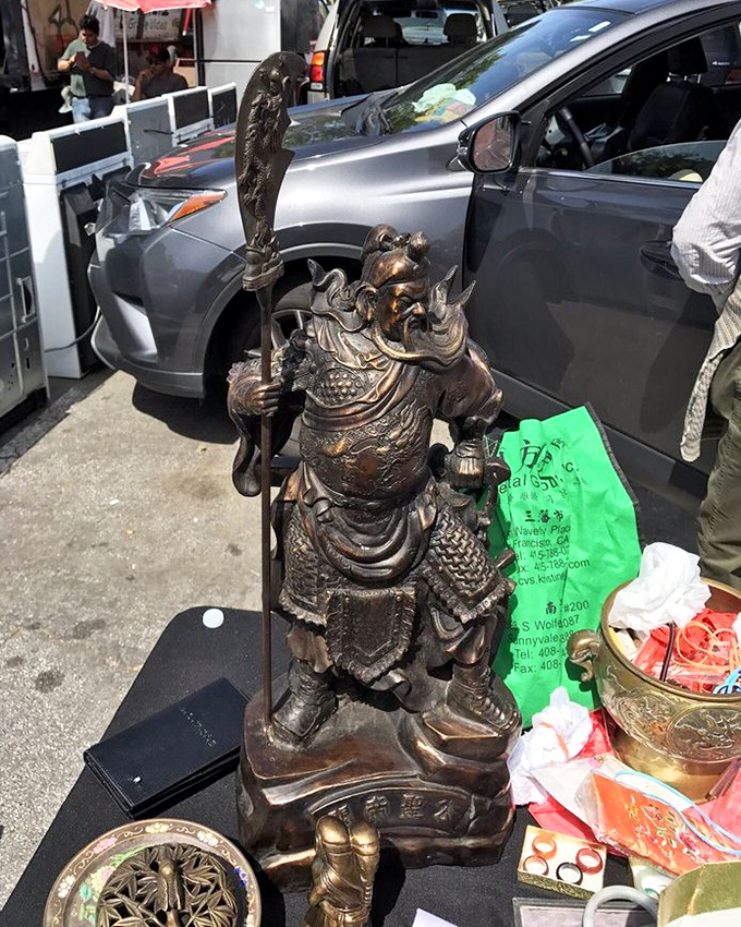 An ornate warrior statue stands guard over treasures unknown. This bronze sentinel has journeyed far to find himself on a folding table in Oakland.