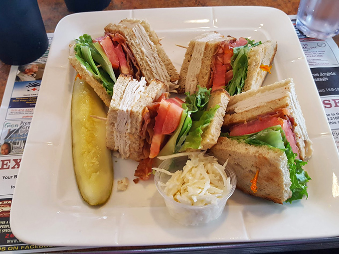 A club sandwich stacked higher than most people's weekend ambitions. The pickle isn't just garnish—it's necessary acid balance.