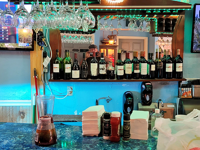 The bar glows with aqua light, bottles standing at attention like soldiers guarding liquid treasures. Wine glasses hang overhead like crystal jellyfish.