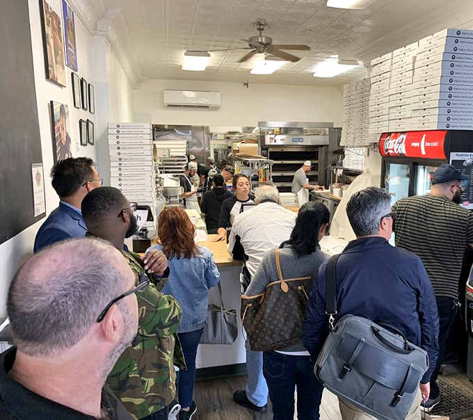 Inside Angelo's, the organized chaos of lunchtime rush is a beautiful dance. Pizza boxes stacked high like edible skyscrapers while hungry patrons wait their turn.