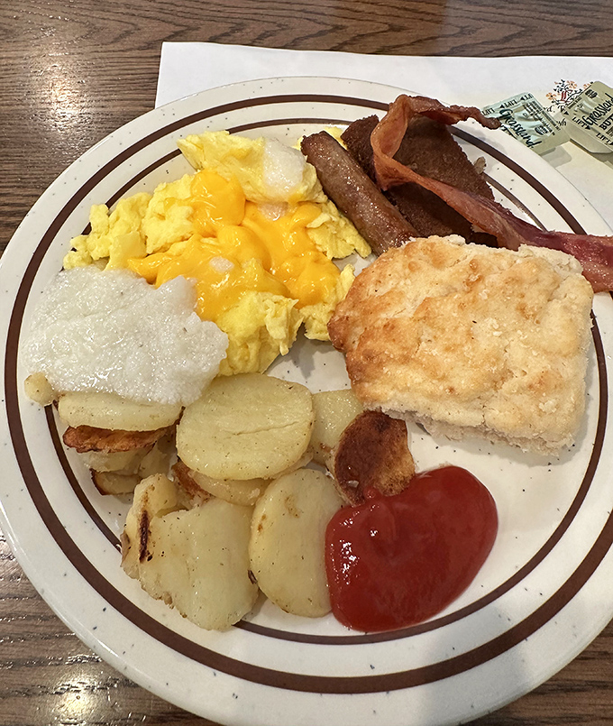 Breakfast perfection on a plate: cheese-topped scrambled eggs, crispy bacon, home fries, and a biscuit that could make your grandmother jealous.