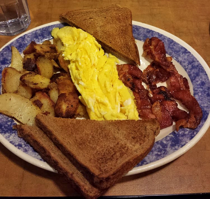 Breakfast perfection on a blue-rimmed plate: fluffy scrambled eggs, crispy home fries, and bacon that strikes that magical balance between crisp and chewy.