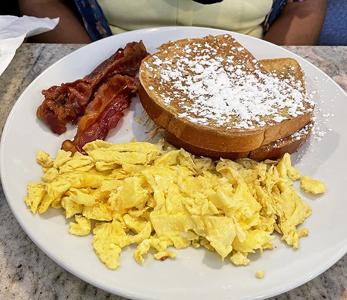 Breakfast nirvana exists, and it looks like this plate: golden French toast dusted with powdered sugar, perfectly scrambled eggs, and bacon that would make vegetarians question their life choices.
