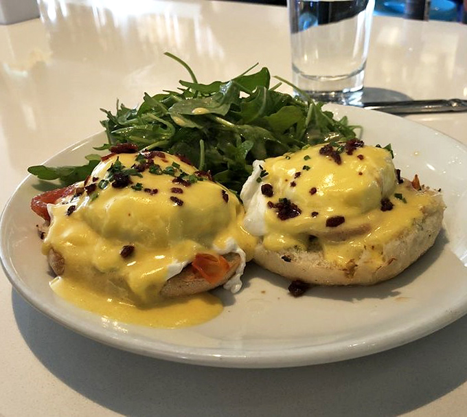 Eggs Benedict perfection: Two golden-yolked masterpieces draped in sunshine-yellow hollandaise with a side of greens pretending this is a balanced meal.