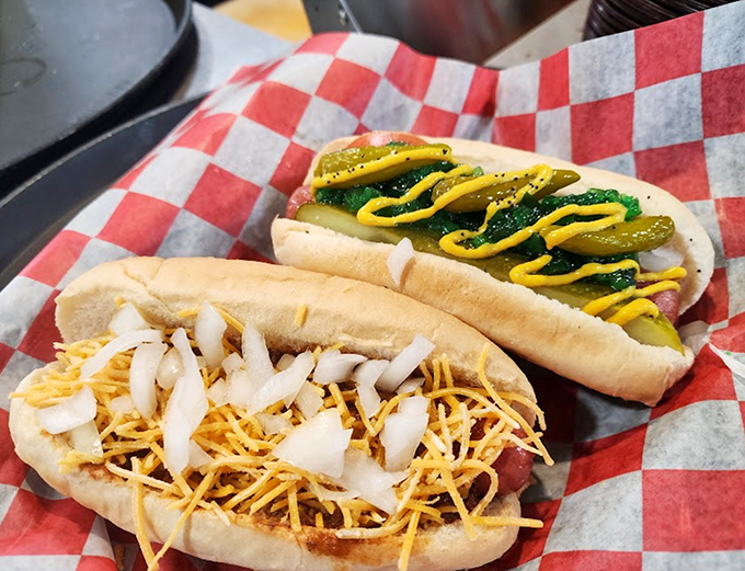 Two dogs living their best life &ndash; one crowned with pickle and mustard, the other buried under cheese and onions. This is hot dog royalty in its natural habitat.