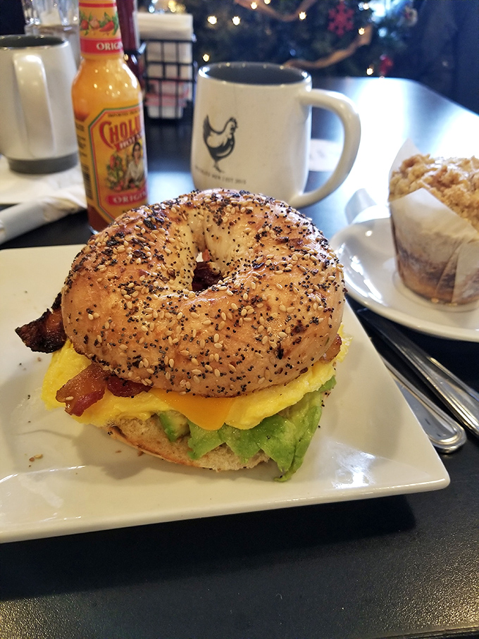 Breakfast sandwich architecture at its finest—a perfectly seeded bagel cradling avocado, egg, and bacon, with that signature Speckled Hen mug standing guard.