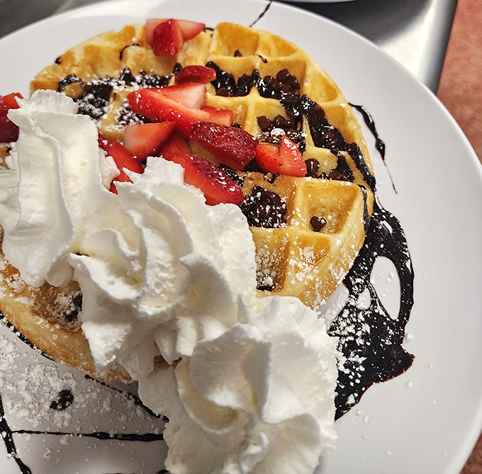 Breakfast nirvana achieved: a golden waffle crowned with fresh strawberries, chocolate drizzle, and whipped cream mountains that would make Mount Mitchell jealous.