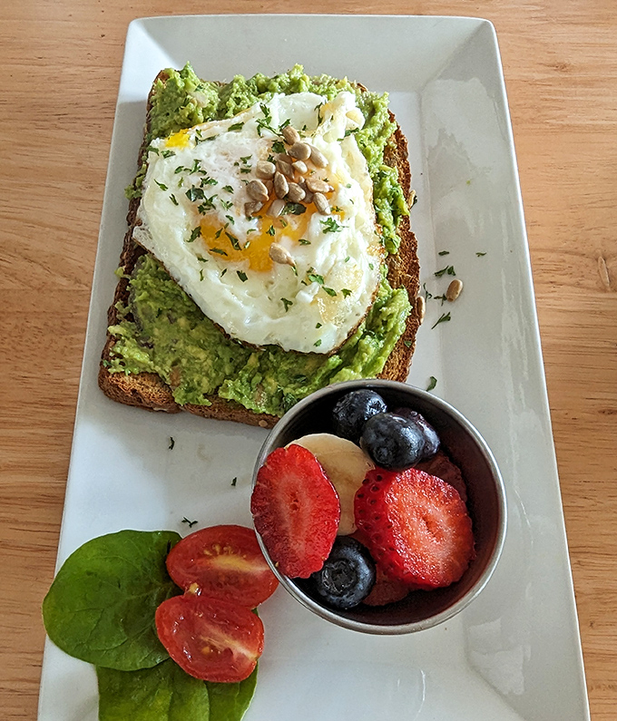 Avocado toast elevated to art form: creamy green goodness topped with a perfectly poached egg, sunflower seeds, and a side of fresh fruit that makes "eating healthy" feel downright indulgent.