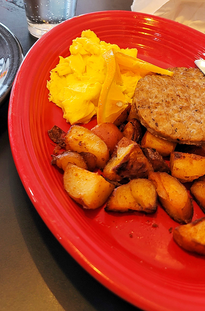 Breakfast perfection on a fiery red plate: golden home fries with crispy edges, fluffy scrambled eggs, and a sausage patty that means business.