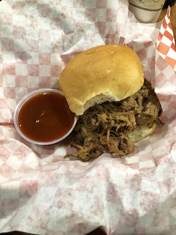 A pulled pork sandwich so tender it practically surrenders to gravity. That sauce cup isn't optional&mdash;it's your ticket to flavor paradise.