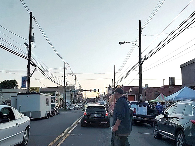 A lively street scene in Nanticoke captures everyday life, where cars and pedestrians navigate a main road lined with utility poles, shops, and a pop-up market.