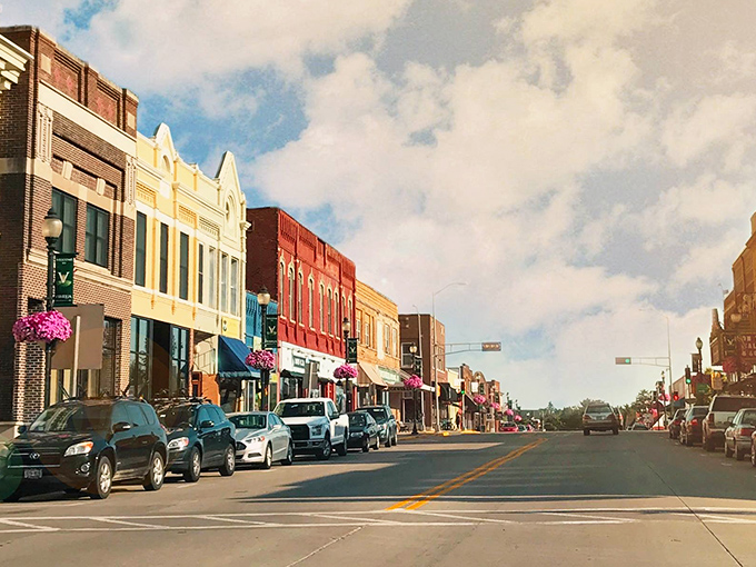 Colorful historic storefronts line a vibrant main street, with hanging flower baskets adding splashes of purple against a backdrop of puffy summer clouds.