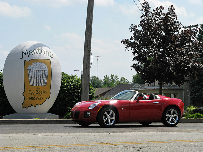 The sleek sports car provides perfect scale comparison to Mentone's oversized egg &ndash; speed meets whimsy at this Indiana intersection.
