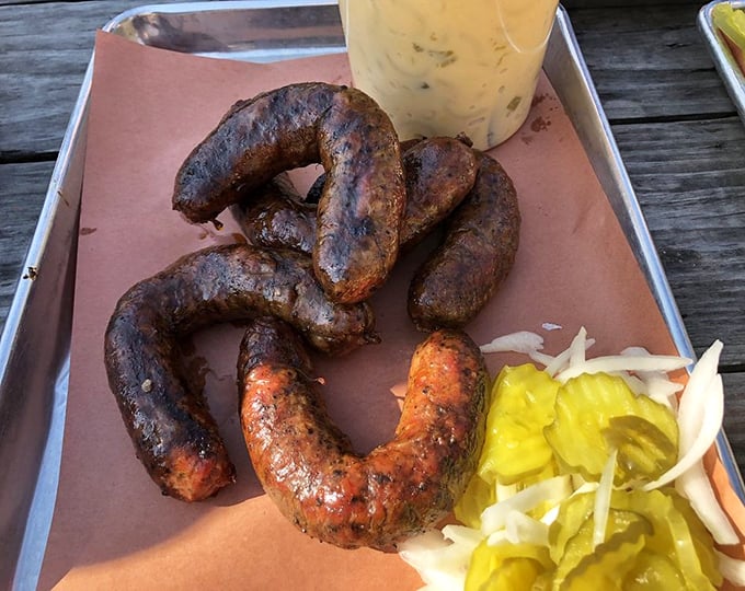 Sausage links curved like smiles, knowing they're about to make your taste buds dance. The pickle garnish isn't decoration&mdash;it's necessary balance.