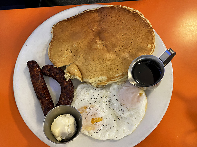 The holy trinity of breakfast: golden pancake, perfectly fried egg, and sausage links that snap when you bite them. Simple perfection on a white plate.