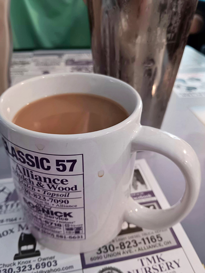 That classic white mug isn't just holding coffee—it's cradling liquid optimism, served against a backdrop of local business ads that tell Alliance's story.